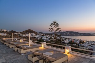 Terraço com espreguiçadeiras e mesas, com vista para uma cidade costeira de edifícios brancos e uma vista clara do mar.
