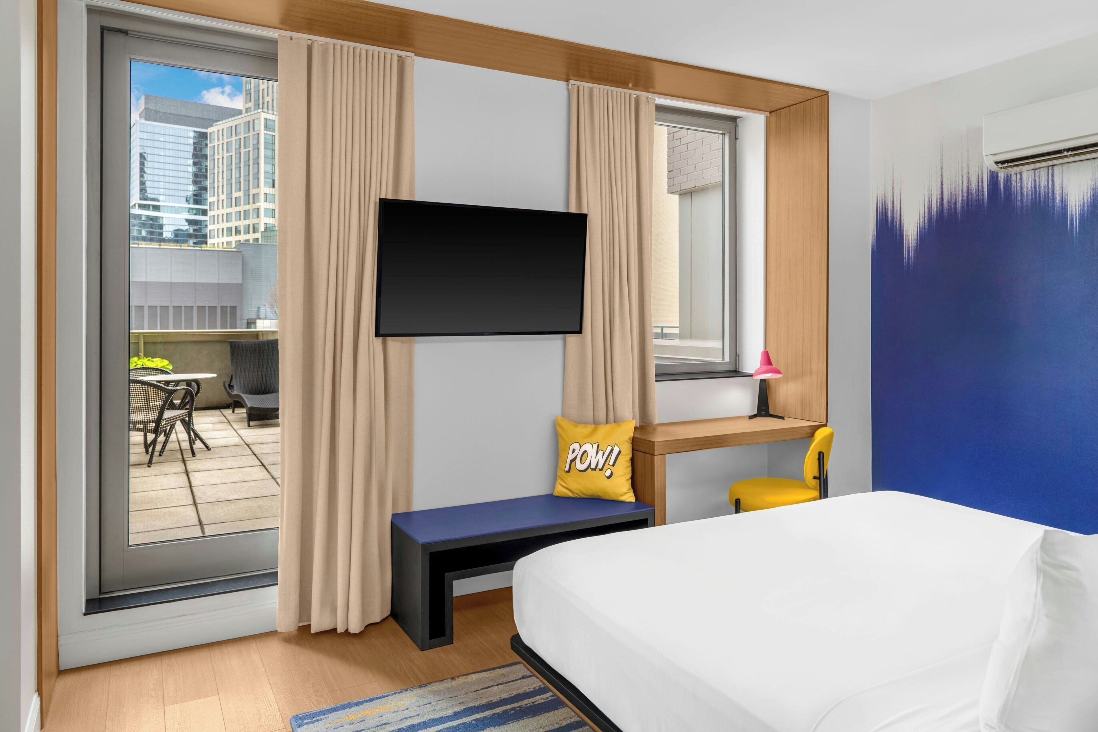Quarto de hotel moderno com uma cama grande, televisão de tela plana na parede, um banco, uma pequena escrivaninha com cadeira e vista da cidade através de amplas janelas.
