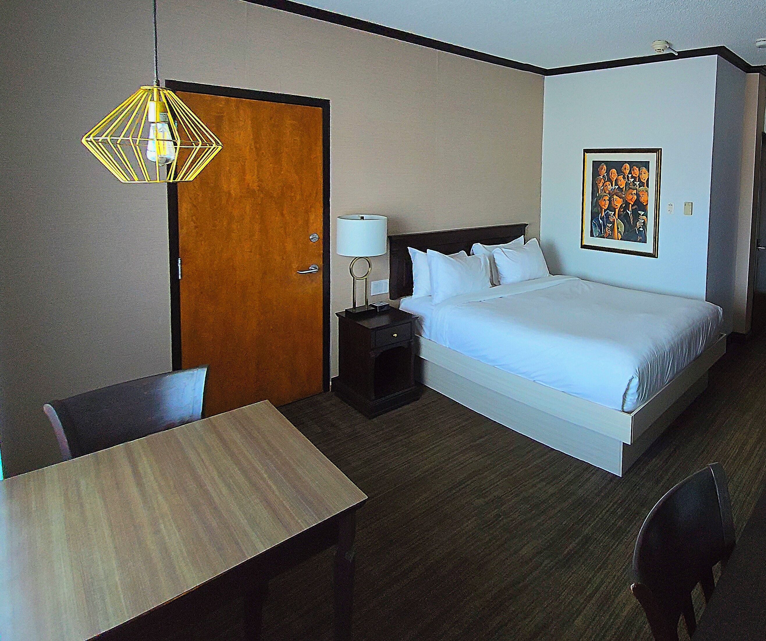Quarto de hotel com cama, mesa de cabeceira, luminária, cadeira e um quadro na parede.
