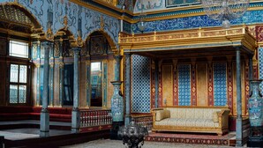 Um quarto luxuoso com desenhos complexos em azul e dourado nas paredes, um grande lustre ornamentado e um sofá macio.