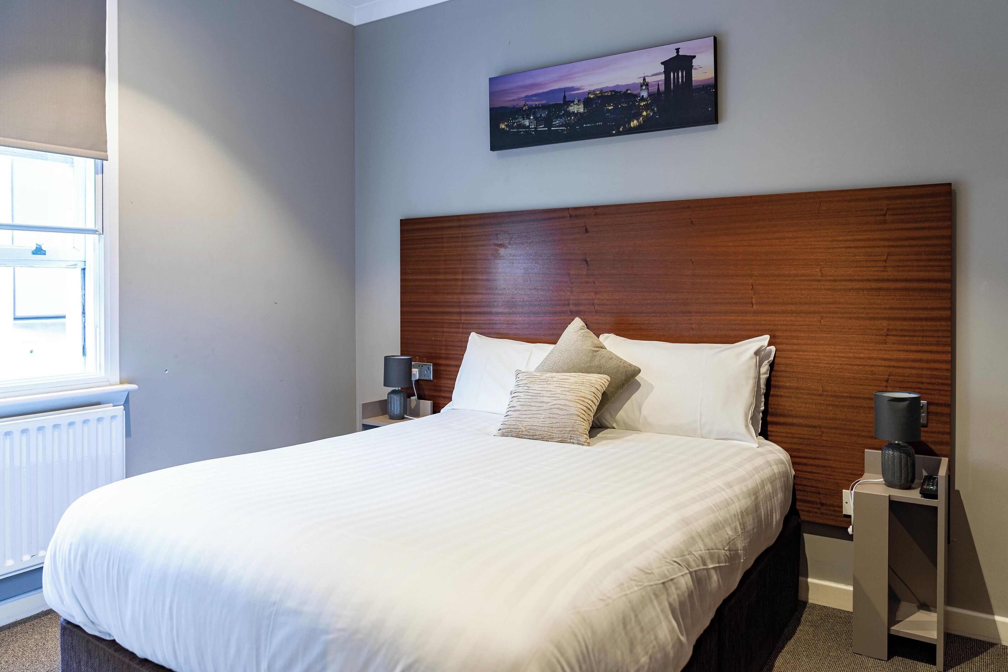 Quarto de hotel com uma cama grande, cabeceira de madeira, duas mesas de cabeceira com abajures, uma janela com persianas e um quadro com vista para a cidade na parede.