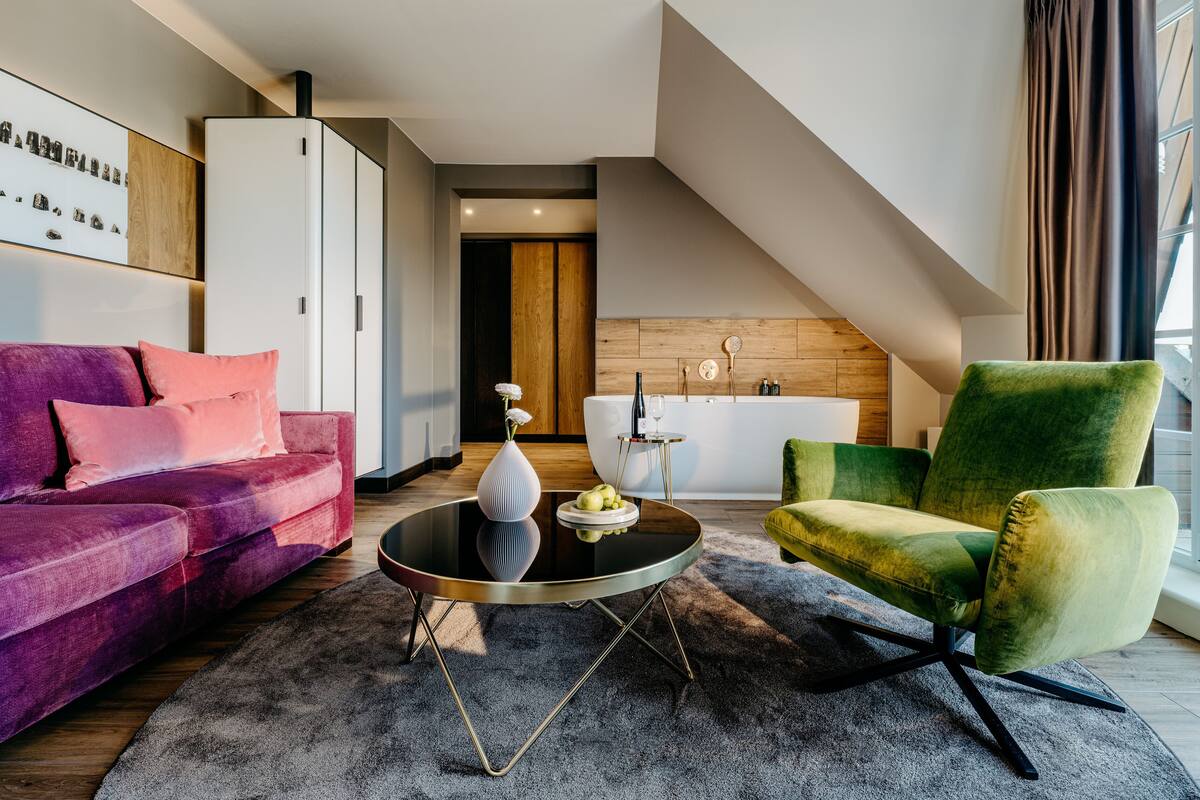 Sala de estar moderna com um sofá roxo, uma poltrona verde, uma mesa de centro redonda e uma área de cozinha ao fundo.
