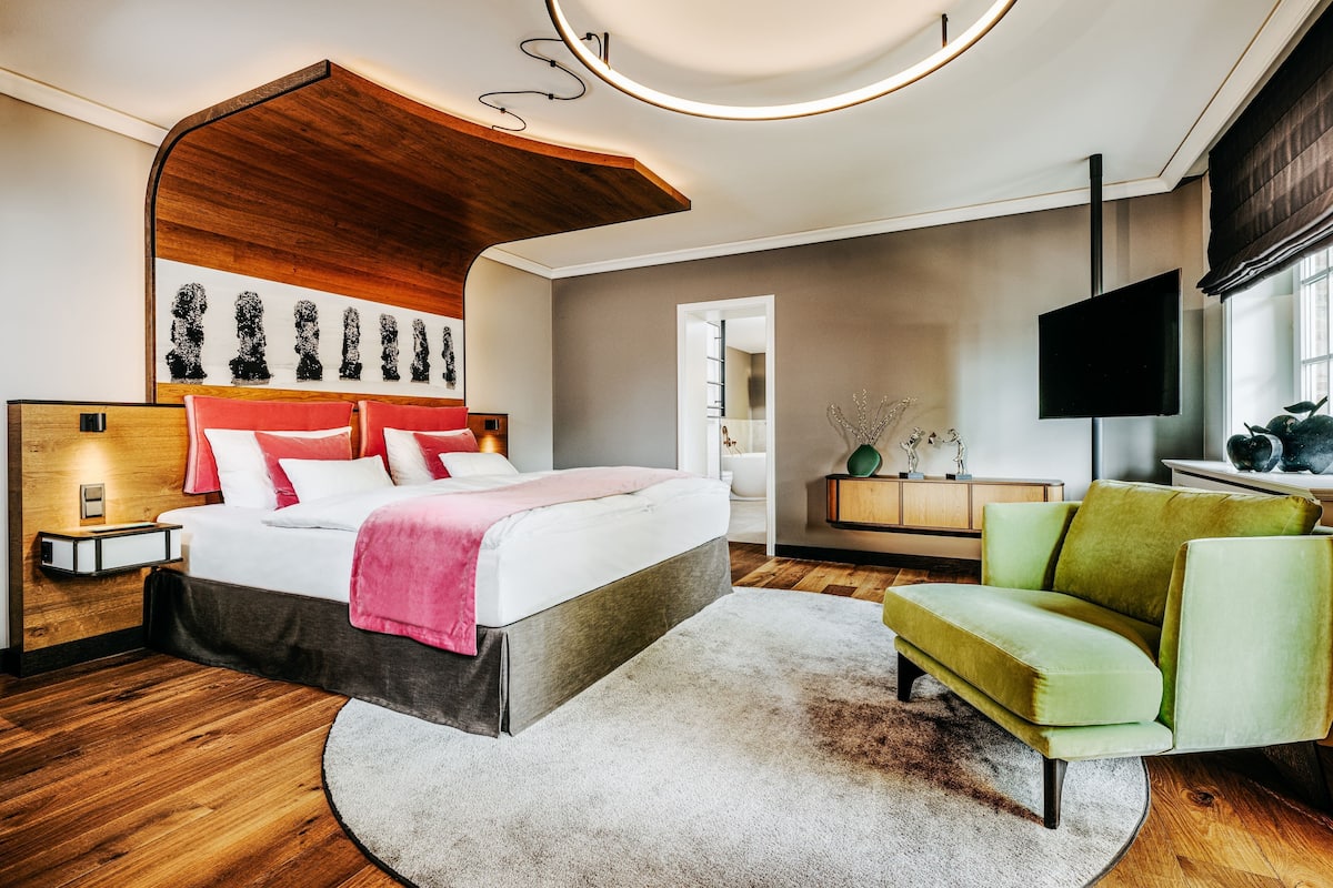 Quarto moderno com teto de madeira, cama com roupa de cama rosa, poltrona verde e televisão de tela plana.