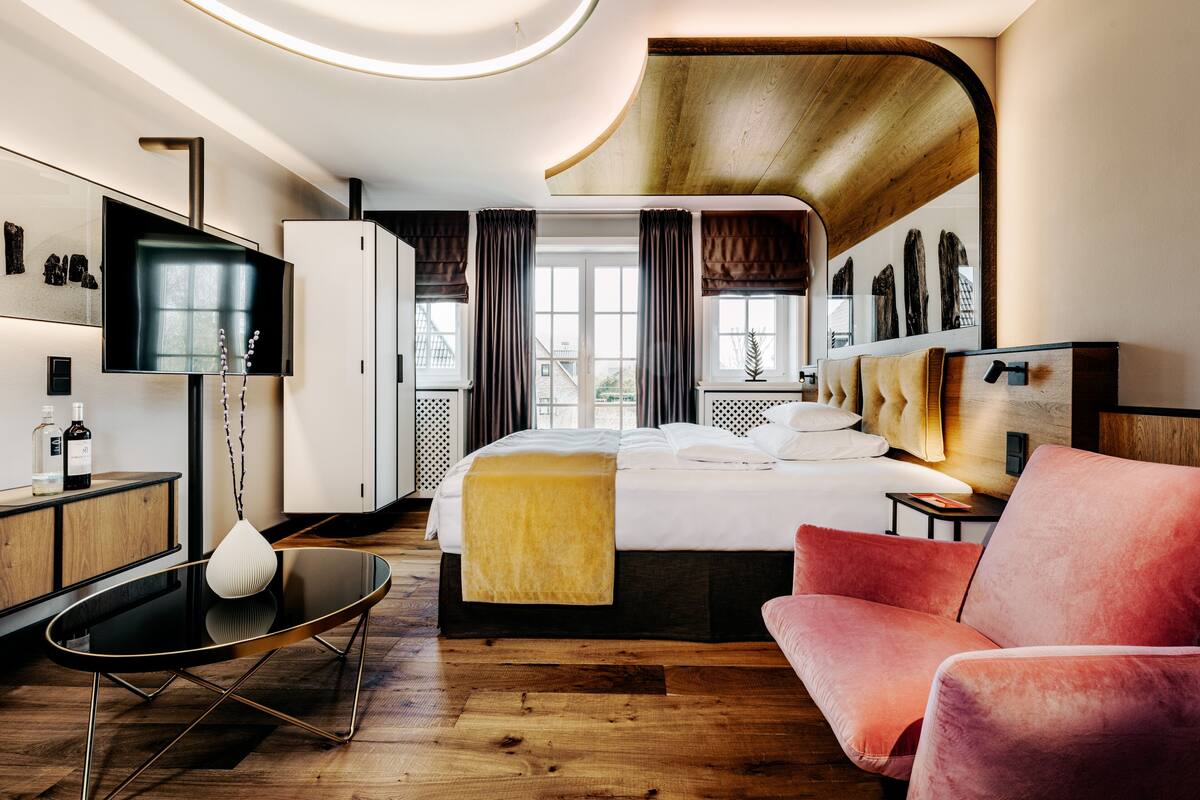 Quarto de hotel moderno com uma cama, um sofá rosa, uma mesa de centro preta, uma televisão e teto de madeira.