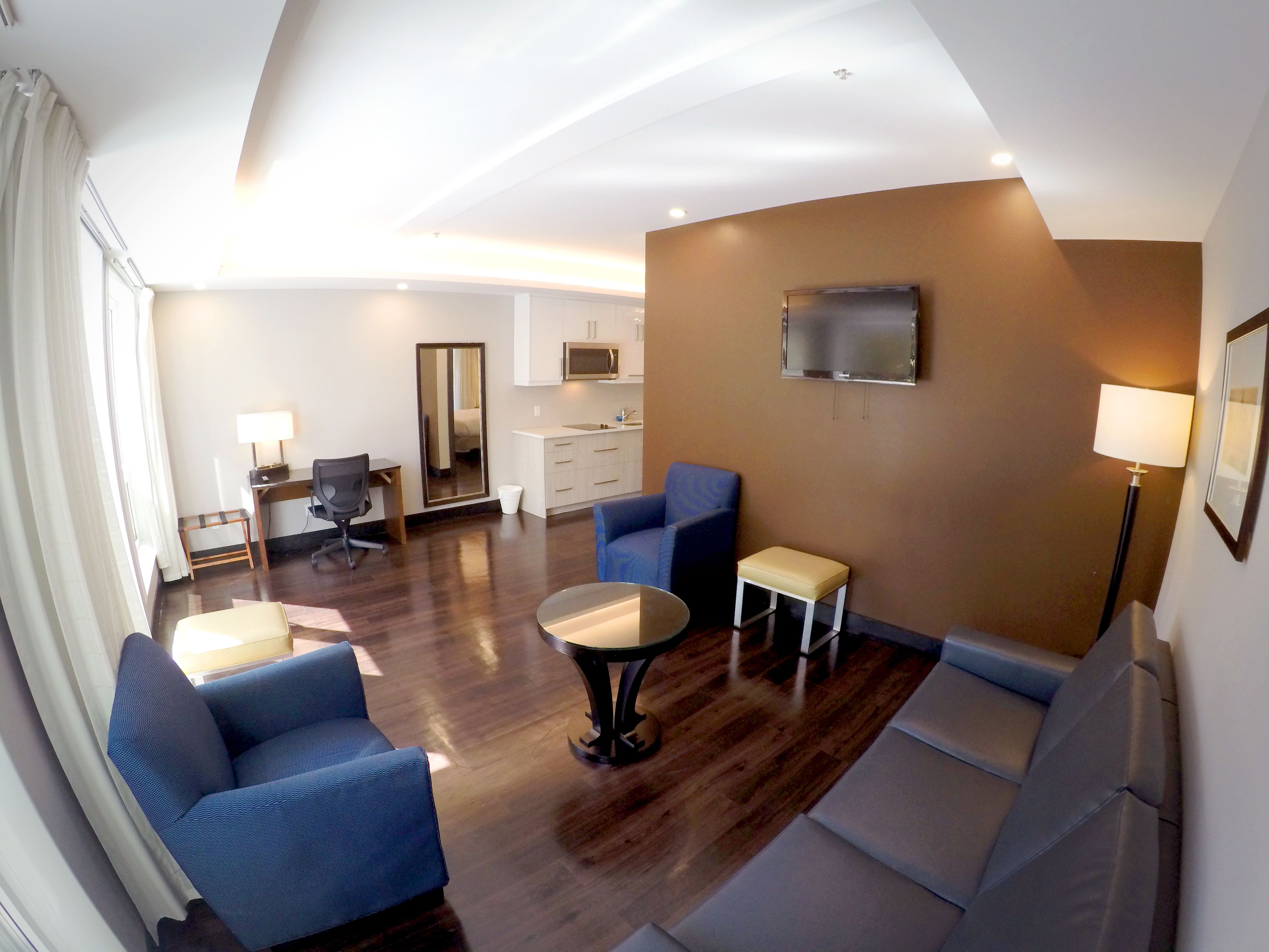 Quarto de hotel moderno com piso de madeira, área de estar com sofás azuis e uma pequena mesa redonda, cozinha compacta com micro-ondas e um espelho grande.
