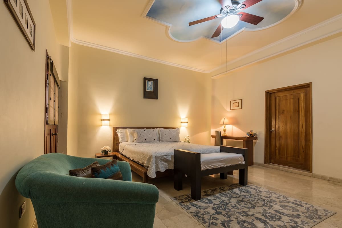 Quarto com cama, duas luminárias de cabeceira, um ventilador de teto, uma porta de madeira e uma poltrona verde.