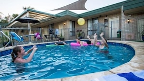Piscina com área de terraço coberta, espreguiçadeiras e um edifício com telhado em azulejos.