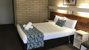 Quarto de hotel com parede em padrão tijolo, cabeceira de madeira, cama bem arrumada com roupa de cama branca e mesa de cabeceira pequena com abajur.