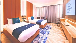 Quarto de hotel com duas camas, uma escrivaninha e uma televisão exibindo o nome de um hotel.
