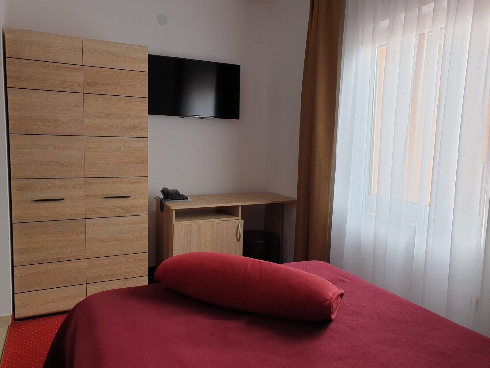 Quarto com guarda-roupa de madeira, televisão de tela plana, secretária com telefone, cama vermelha e cortinas leves.