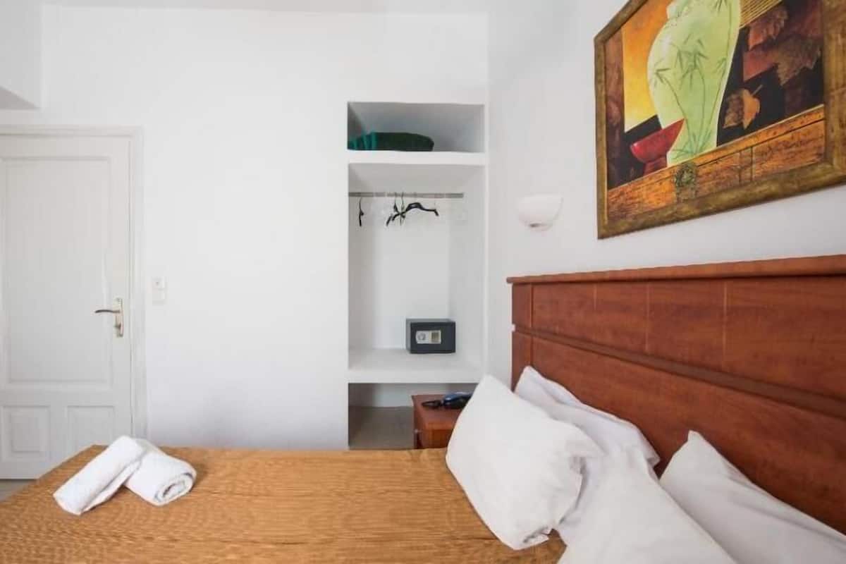 Quarto de hotel com cama de madeira, almofadas brancas, toalha enrolada, pequena mesa de cabeceira, prateleira com relógio, cabideiro e um quadro na parede.