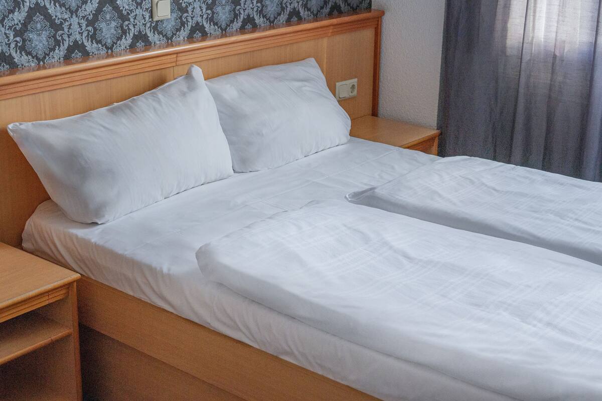 Uma cama de casal com lençóis brancos, cabeceira de madeira e mesa de cabeceira com abajur.