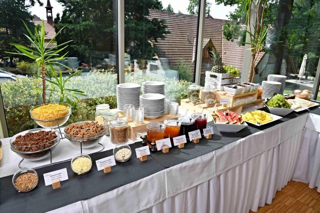 Mesa de buffet com diversos alimentos e bebidas, rodeada por janelas de vidro com vista para árvores e edifícios.