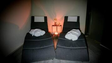Duas camas de solteiro com cabeceiras escuras e roupa de cama branca, uma mesa de cabeceira com um abajur e um pequeno objeto decorativo sobre a mesa.