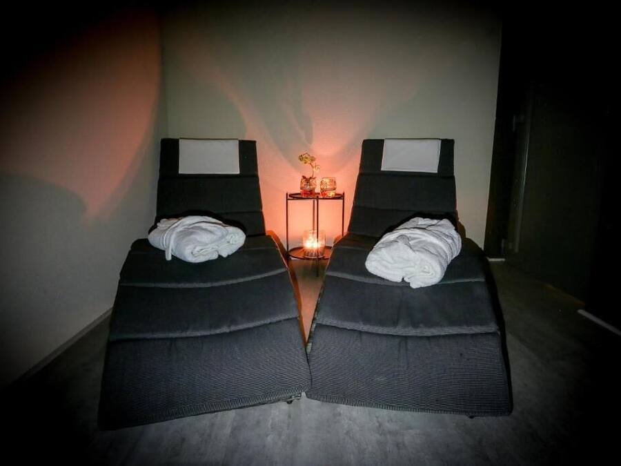 Duas camas de solteiro com cabeceiras escuras e roupa de cama branca, uma mesa de cabeceira com um abajur e um pequeno objeto decorativo sobre a mesa.