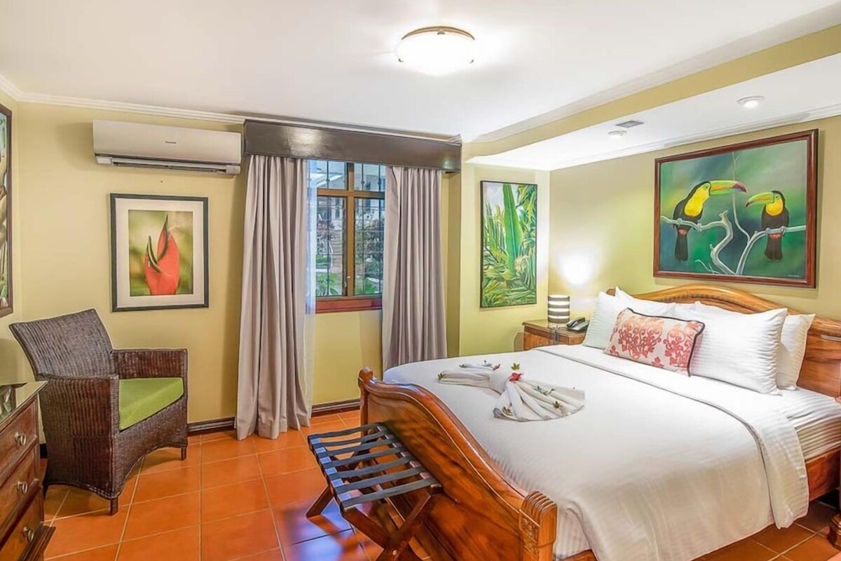 Quarto de hotel com cama, cadeira, janela com cortinas e obras de arte nas paredes.