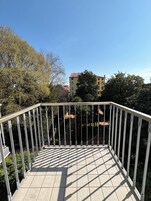 Varanda com chão de azulejos e grades de metal, com vista para um pátio com árvores e um edifício ao fundo.