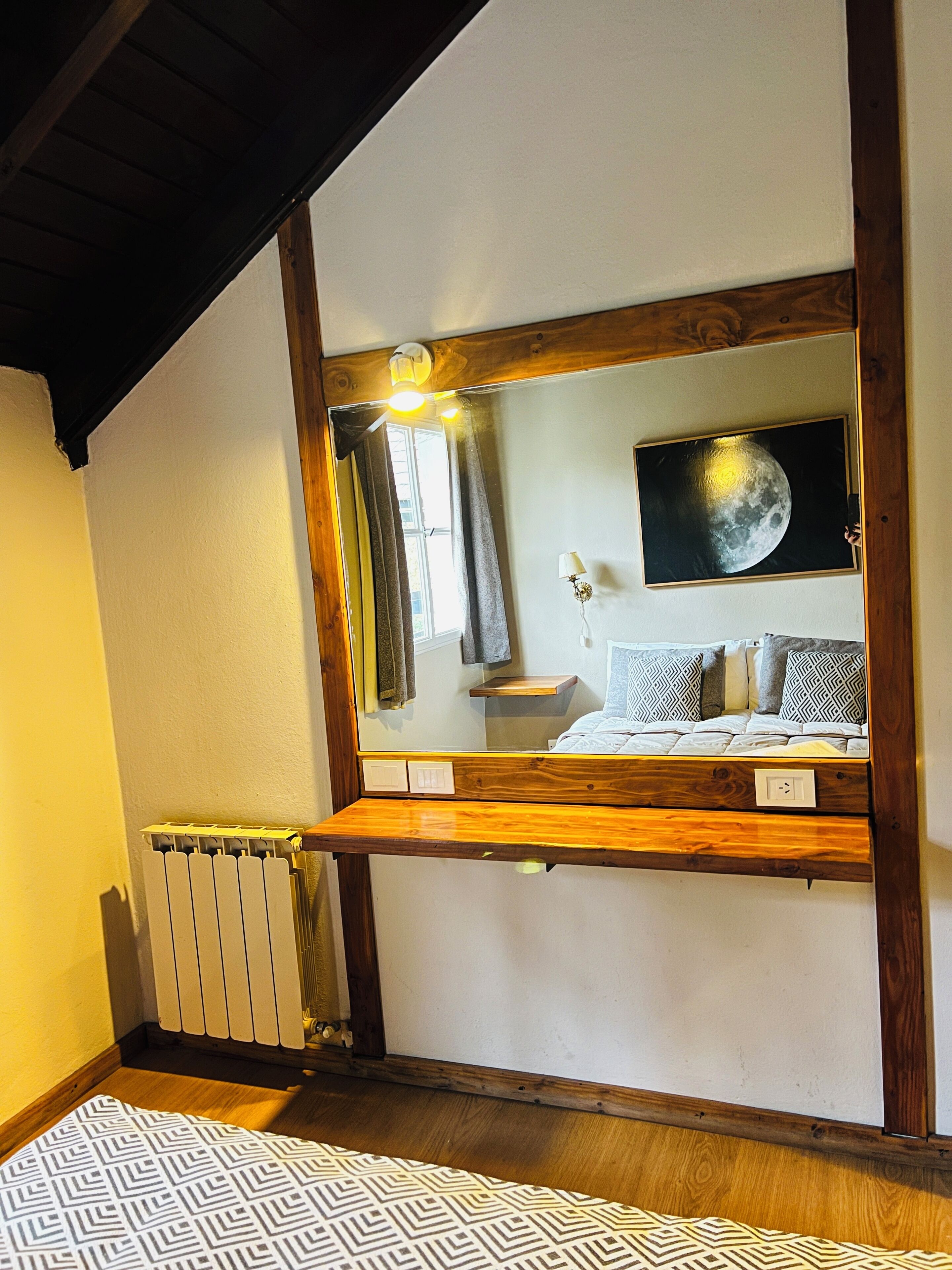 Quarto com teto de madeira, um espelho grande, cama com roupa de cama estampada, mesa de cabeceira e radiador.