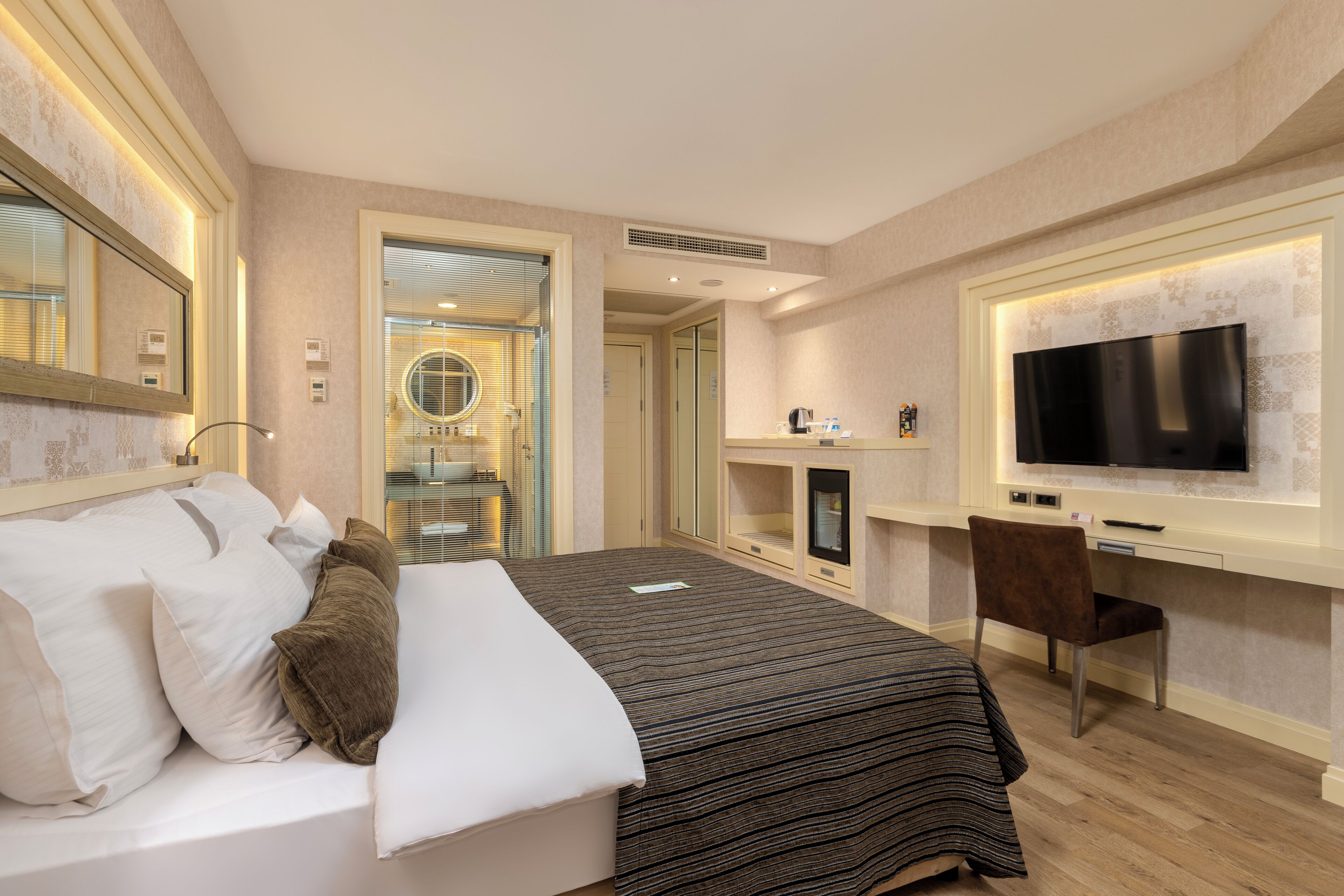 Quarto de hotel moderno com uma cama grande, uma escrivaninha com televisão de tela plana e um banheiro visível através de uma porta de vidro.