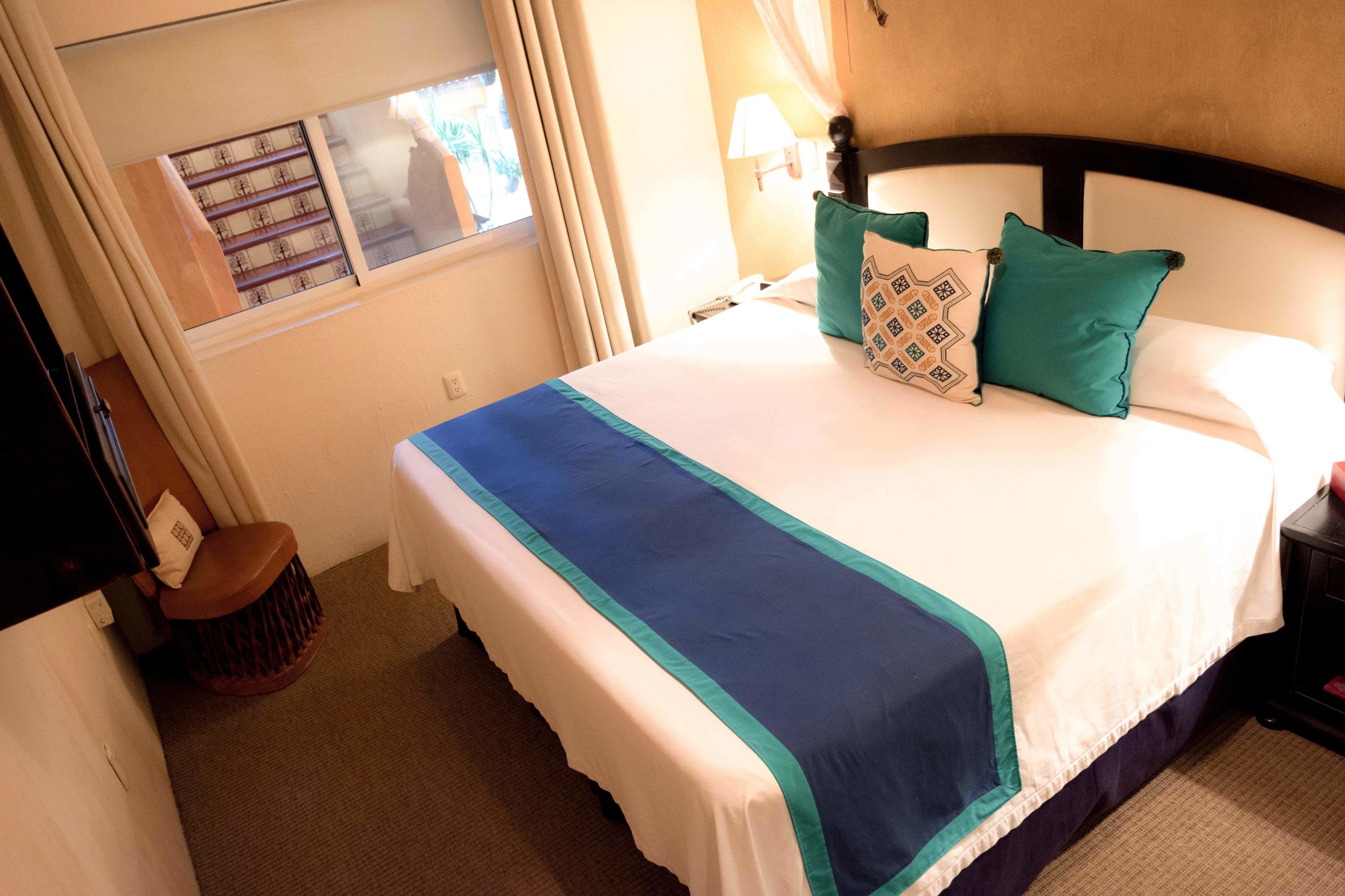 Quarto de hotel com cama, janela com cortinas, mesa de cabeceira e abajur.
