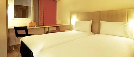 Quarto de hotel com cama de casal, escrivaninha com cadeira, janela com persianas e mesa de cabeceira com luminária.