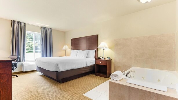 Quarto de hotel com cama, abajures, escrivaninha, janela com cortinas e banheiro com banheira e pia.