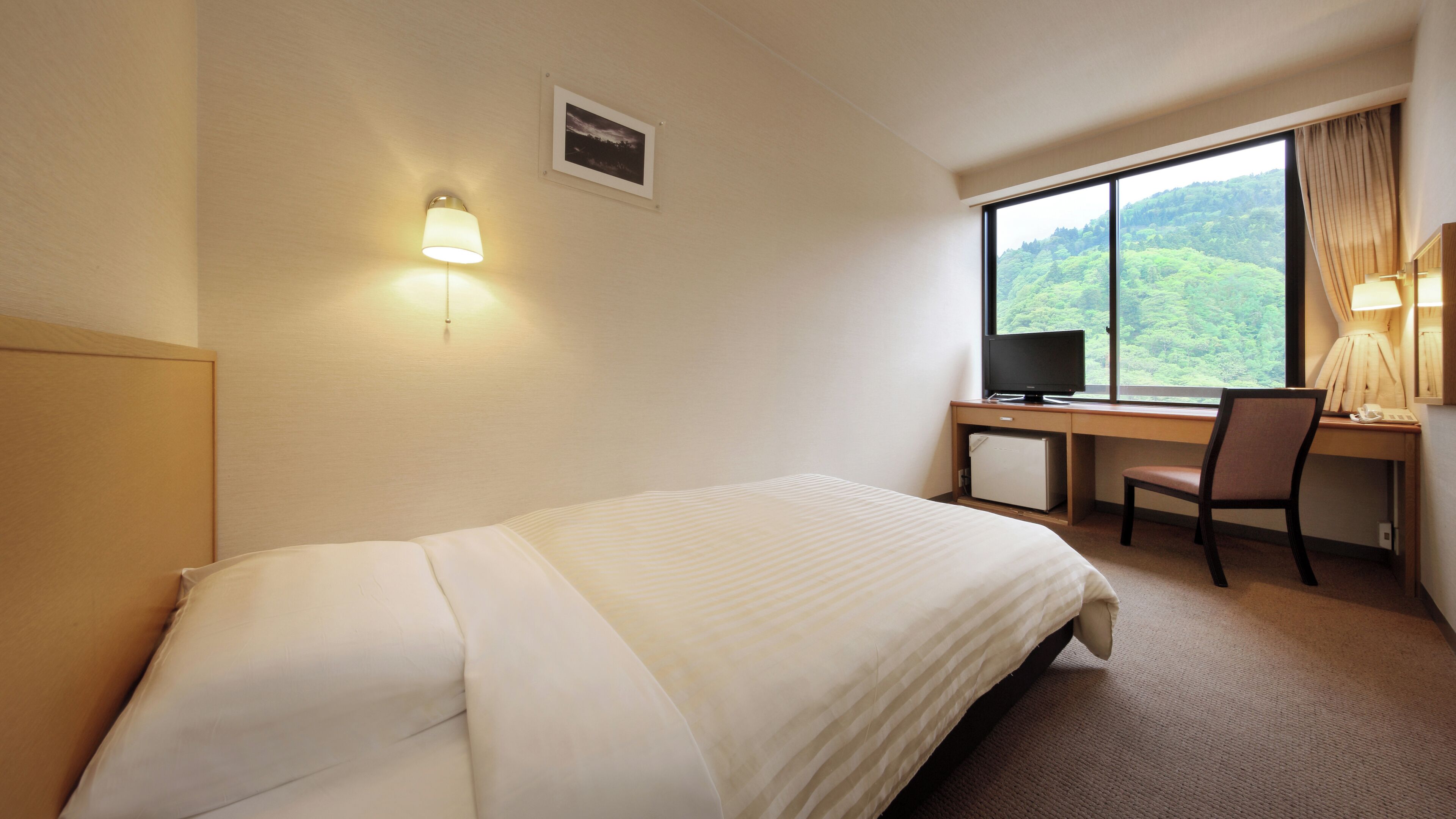 Quarto de hotel com cama, escrivaninha com computador, cadeira, pequena geladeira e janela com vista para uma floresta.