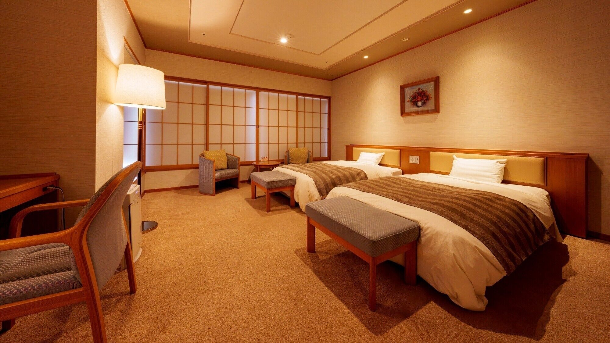Quarto de hotel com porta tradicional japonesa deslizante, cama com cabeceira de madeira, escrivaninha com cadeira e pequena mesa com abajur.