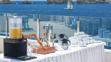 Mesa de buffet com máquinas de café, xícaras e dispensador de suco, com vista para o oceano e um veleiro.