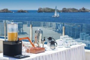 Mesa de buffet com máquinas de café, xícaras e dispensador de suco, com vista para o oceano e um veleiro.