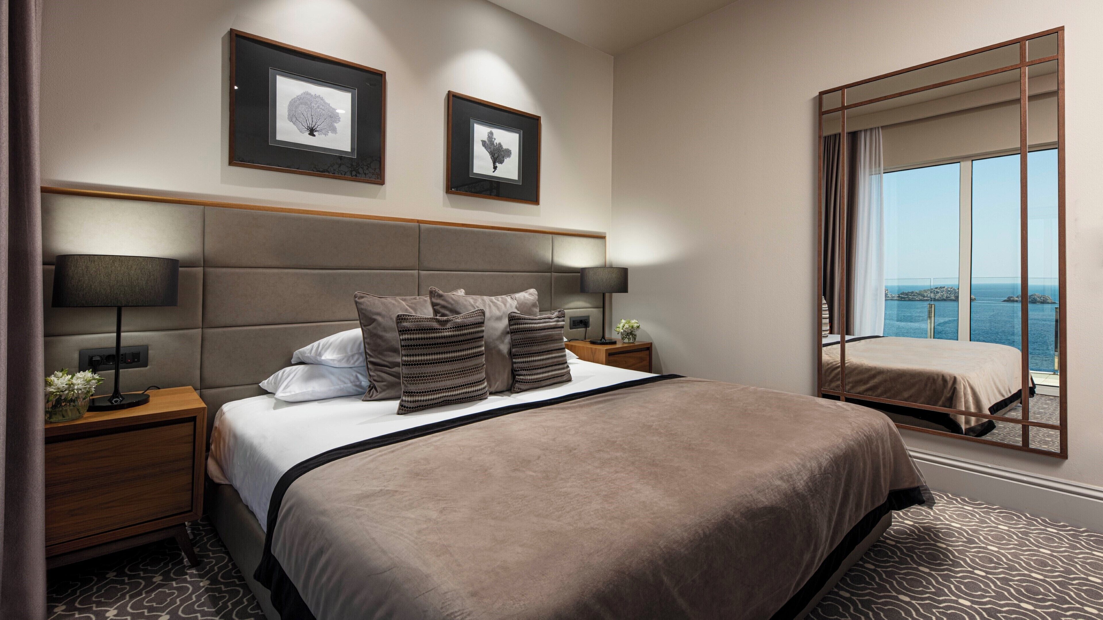 Quarto de hotel moderno com uma cama grande, duas luminárias de cabeceira, uma mesa de cabeceira com abajur e um espelho grande refletindo uma vista panorâmica.