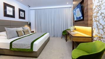 Quarto de hotel com cama, escrivaninha, cadeira, televisão e cortinas.