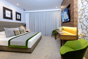 Quarto de hotel com cama, escrivaninha, cadeira, televisão e cortinas.