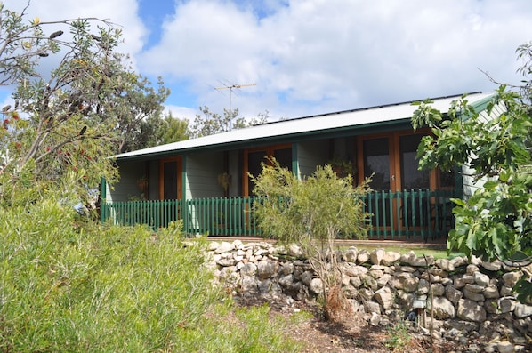 Uma casa de dois andares com varanda verde, muro de pedra e vegetação ao redor.