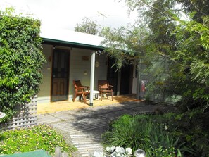 Uma pequena casa com varanda, duas cadeiras de madeira e um jardim com diversas plantas e flores.