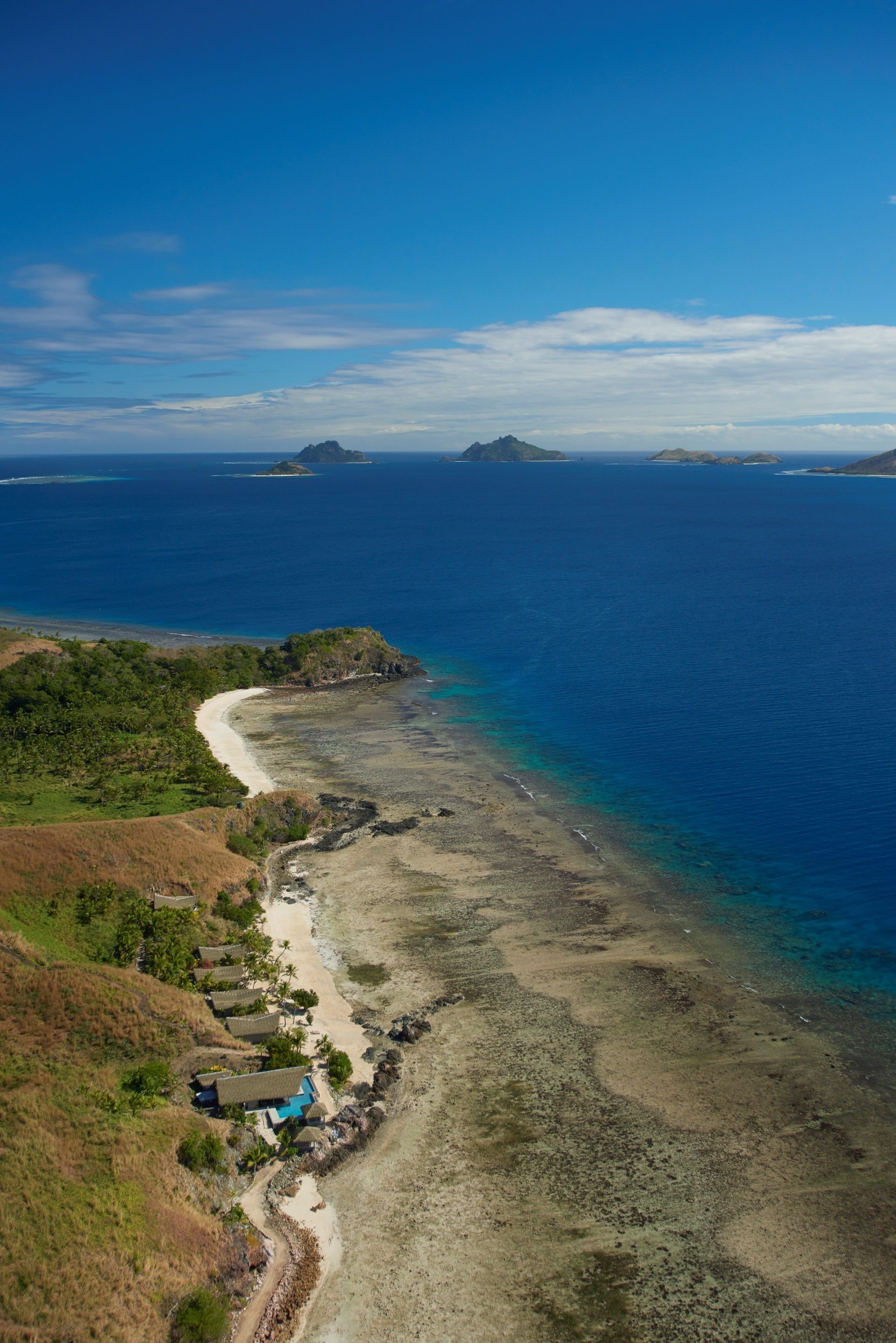 Vista aérea de uma área costeira com uma praia, um pequeno povoado com casas e ilhas ao longe.