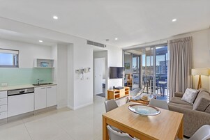 Cozinha moderna com armários brancos, uma pia e uma janela. Há uma sala de jantar com mesa e cadeiras de madeira, e uma sala de estar com sofá e televisão.