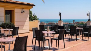 Um terraço ao ar livre com várias mesas e cadeiras redondas, vista para o mar e um edifício com fachada amarela.