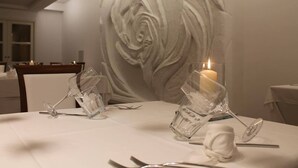 Mesa de jantar posta com copos de vidro transparente e uma vela, com uma grande obra de arte floral na parede ao fundo.