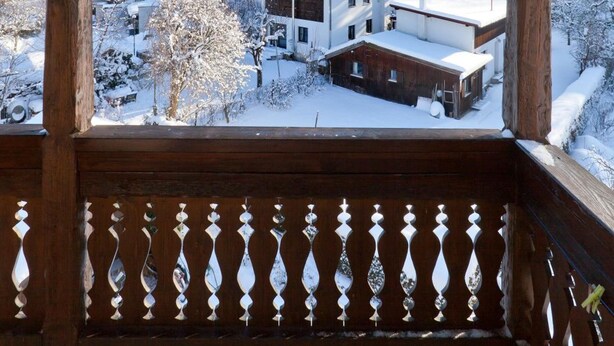 Varanda de madeira com grades ornamentadas, com vista para uma aldeia coberta de neve e um castelo.