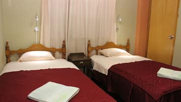Quarto de hotel com duas camas, cada uma com cabeceira de madeira e colcha vermelha. Um criado-mudo entre as camas tem uma luminária.