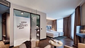 Quarto de hotel moderno com uma cama grande, um sofá, uma mesa de centro e uma TV. Há uma porta de correr com uma frase motivacional.