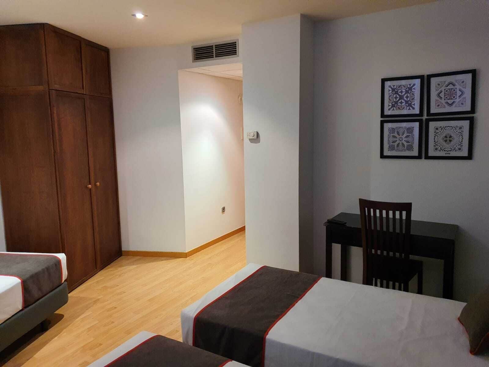 Quarto de hotel com guarda-roupa de madeira, escrivaninha com cadeira e duas camas com detalhes vermelhos.