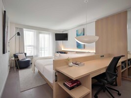 Quarto de hotel moderno com uma cama, uma escrivaninha, uma cadeira, uma TV e uma janela grande com cortinas.