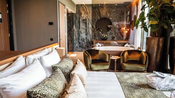 Quarto de hotel moderno com uma cama grande, um sofá, duas poltronas, uma mesinha e um espelho amplo.