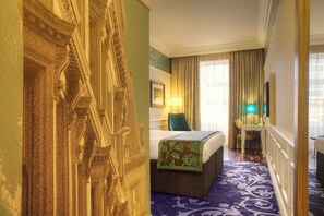 Quarto de hotel com cama, escrivaninha, cadeira e uma janela grande com cortinas.