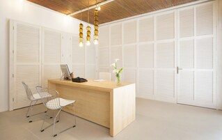 Bancada de recepção com computador, um vaso de flores e uma cadeira em um quarto com armários brancos e teto de madeira.