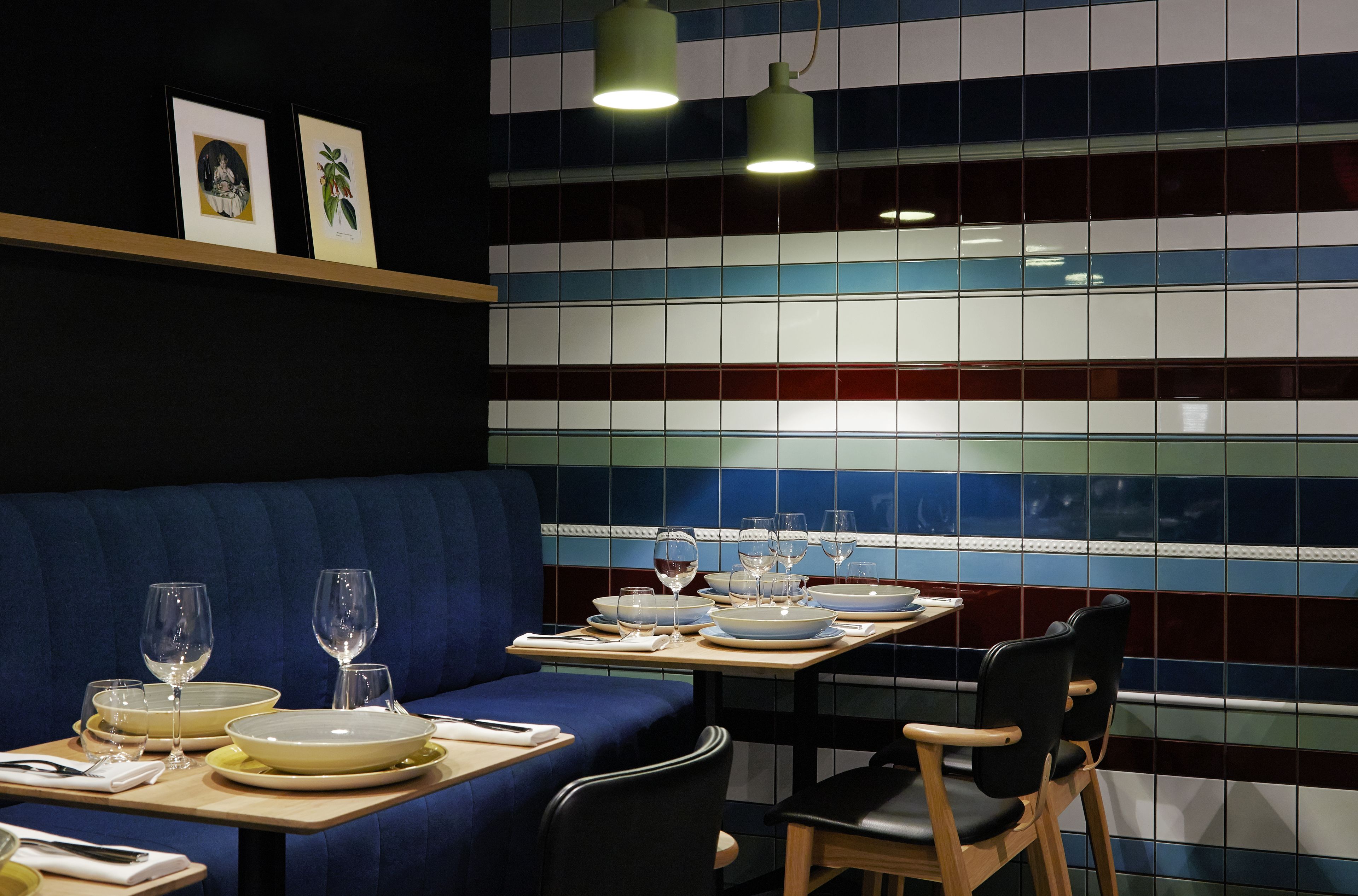 Uma área de refeições com um assento estofado azul, uma mesa posta com pratos, copos e talheres, e uma parede de azulejos com um padrão colorido.
