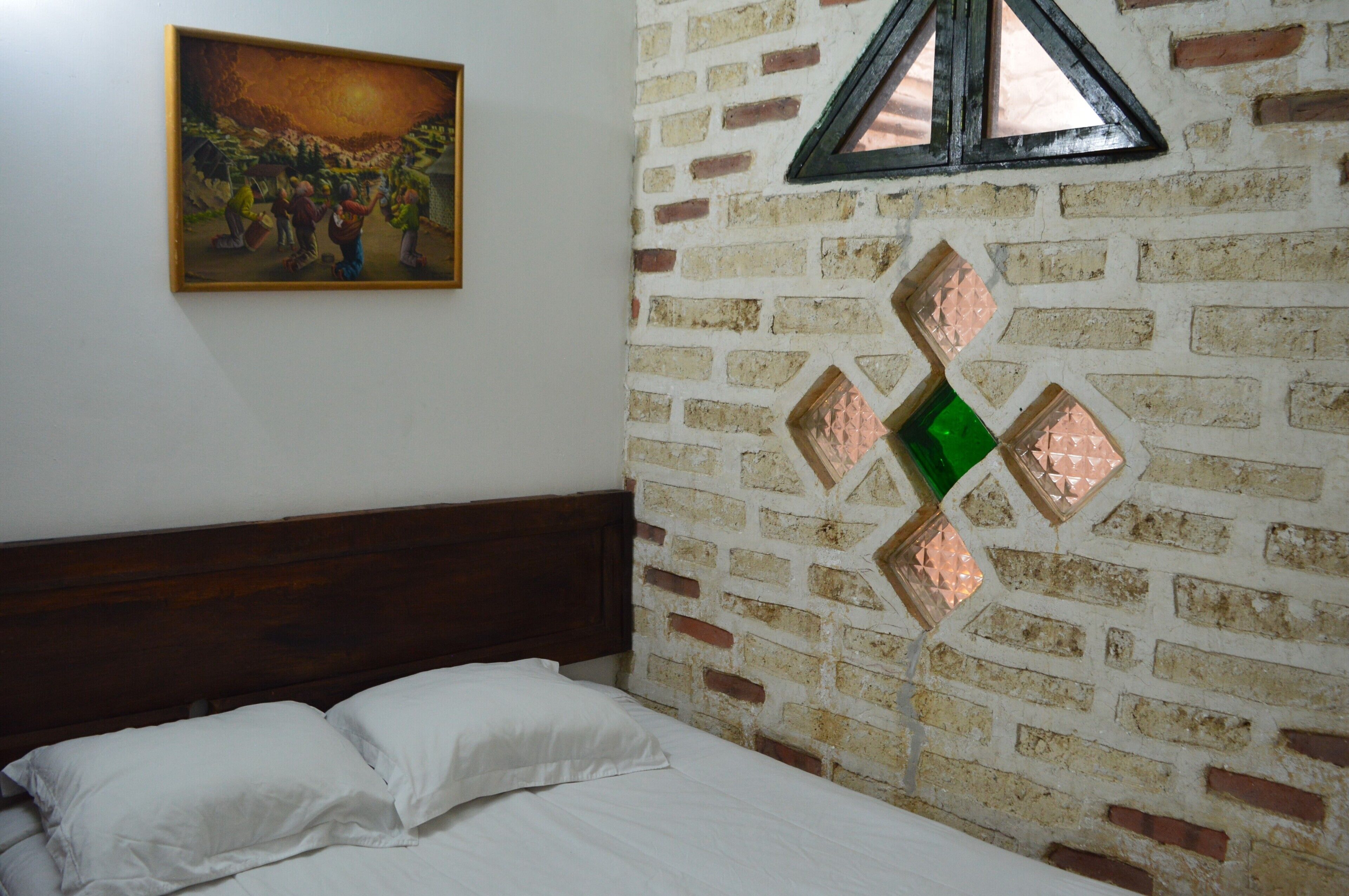 Quarto com cabeceira de madeira, cama com roupa de cama branca, quadro na parede e parede de tijolos com design de janela geométrica.