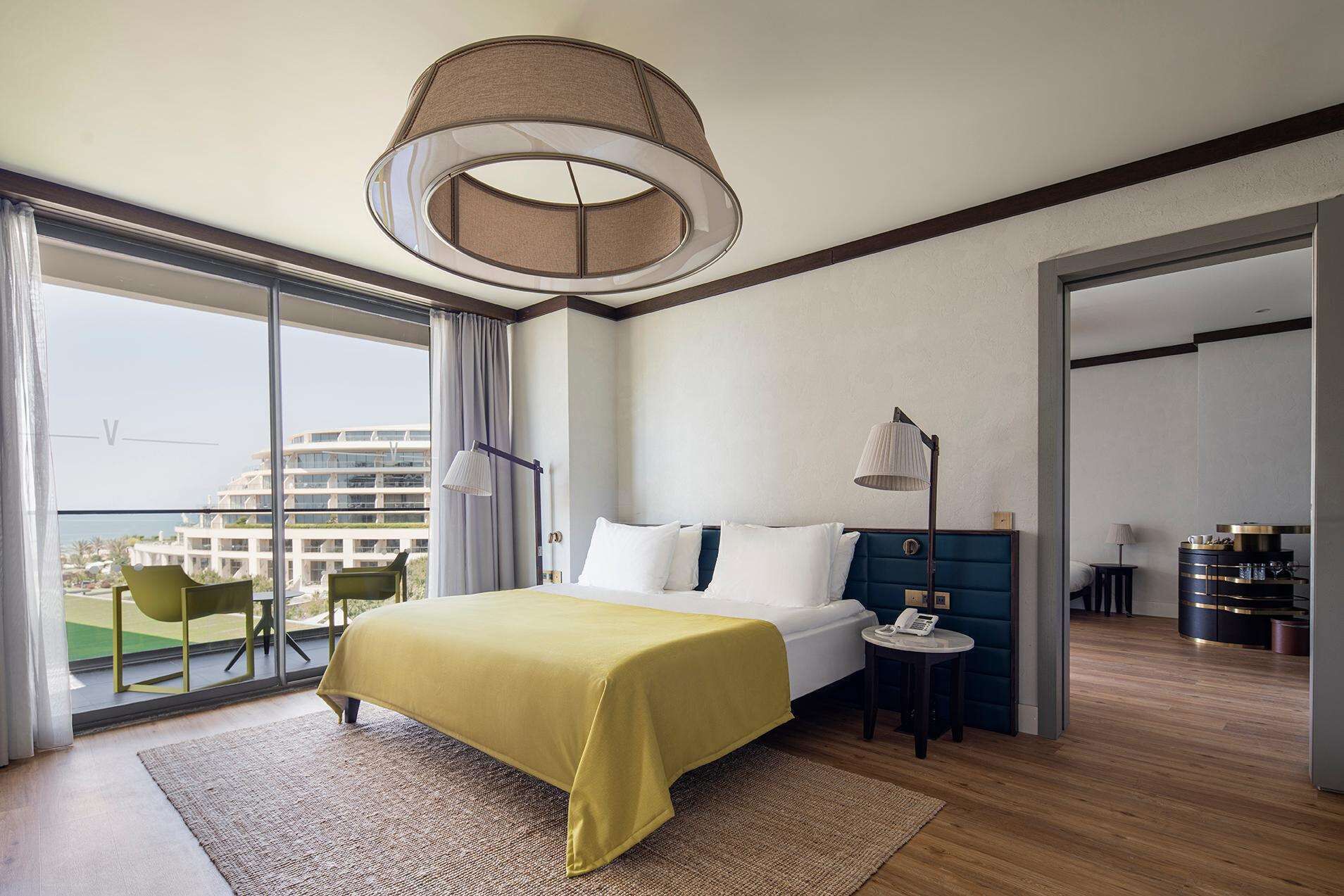 Quarto de hotel moderno com uma cama grande, uma mesinha de cabeceira, uma luminária e vista para a cidade.
